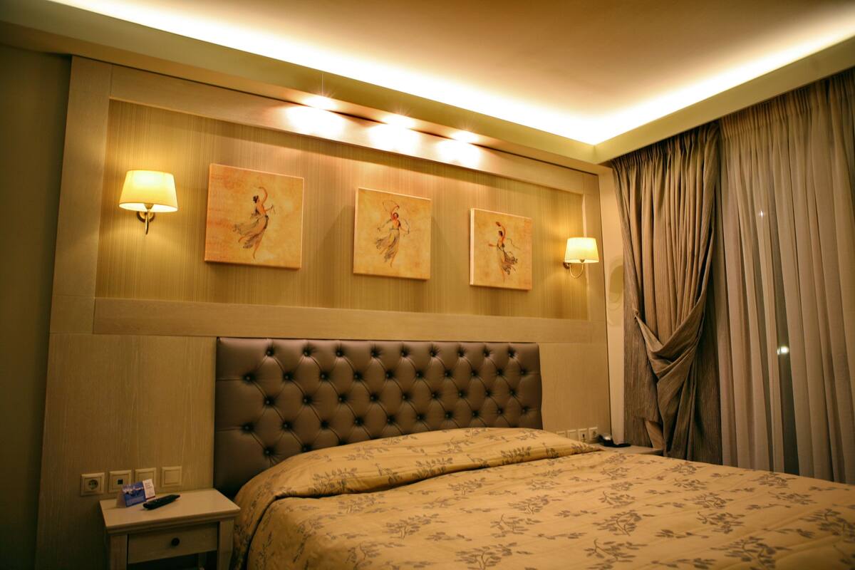 Quarto de hotel com cabeceira estofada, cama com roupa de cama floral, mesa de cabeceira com controle remoto e luminárias fixadas na parede.