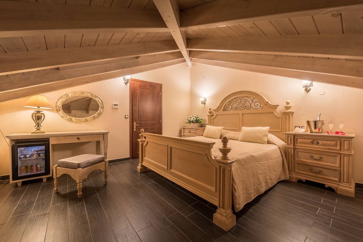 Um quarto com teto de madeira, uma cama com cabeceira, mesa de cabeceira, cómoda, espelho e frigorífico.