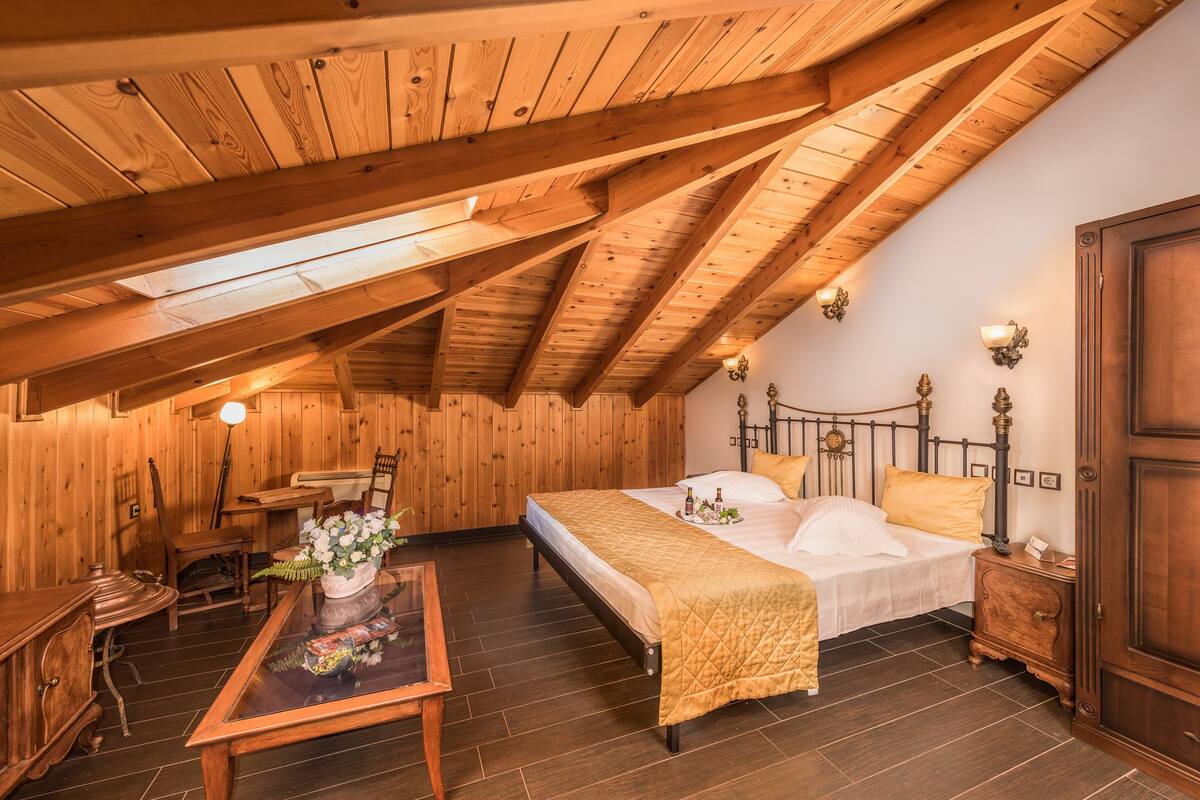 Quarto de madeira com teto inclinado, uma cama com colcha, uma mesa com vaso e uma cadeira.