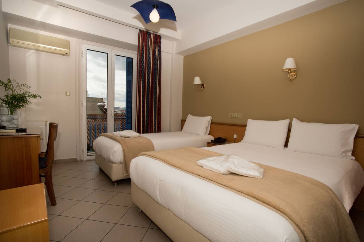 Quarto de hotel com duas camas, uma mesa, uma cadeira, um vaso de plantas e uma varanda com vista.