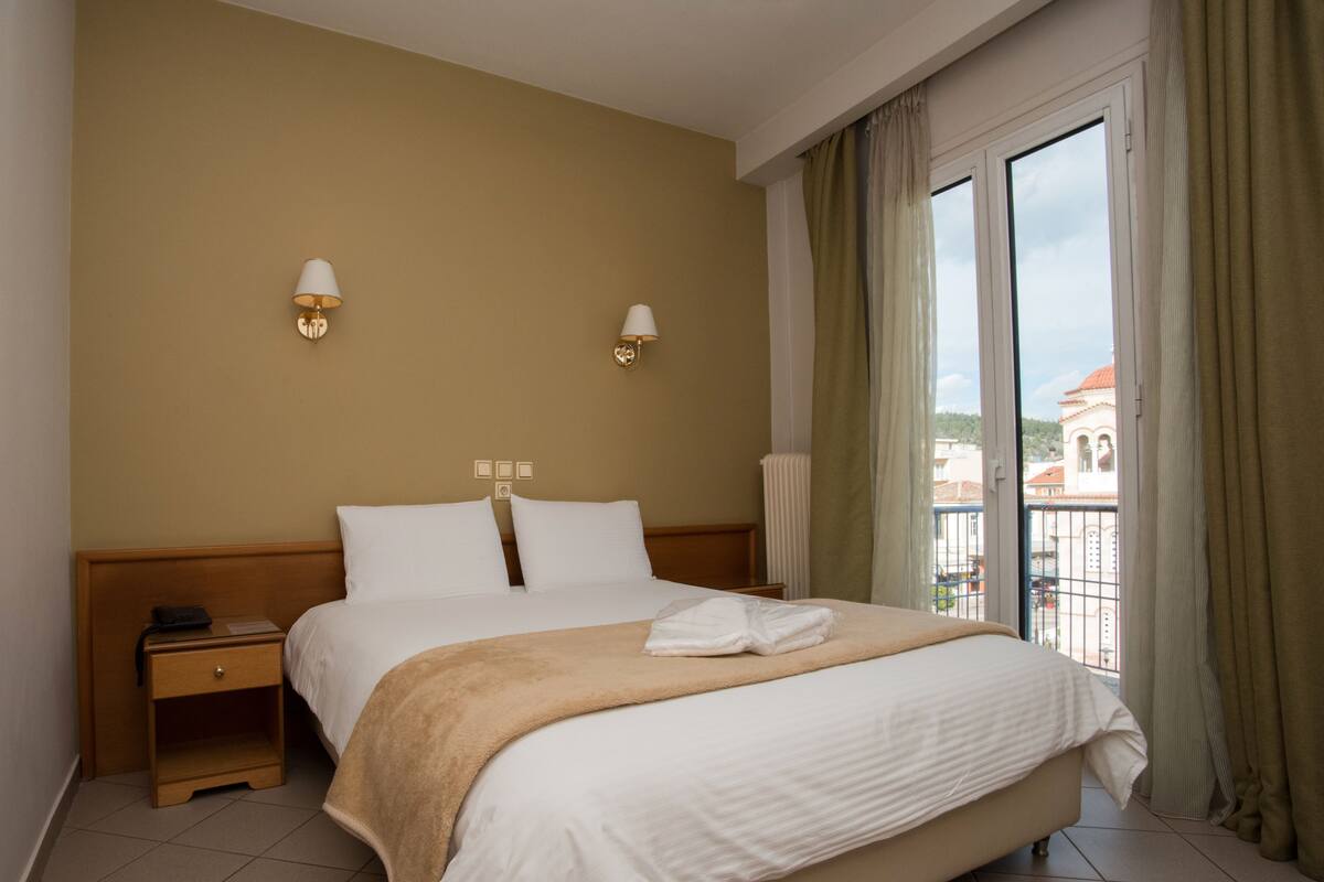 Quarto de hotel com cama, mesa de cabeceira, duas luminárias fixadas na parede e vista de prédios através de uma janela com cortinas.