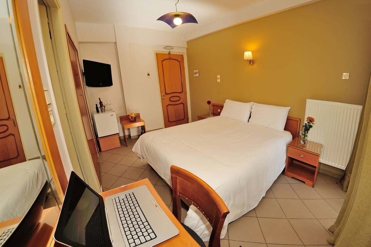 Quarto de hotel com cama, escrivaninha com laptop, mesa de cabeceira com um vaso e televisão na parede.