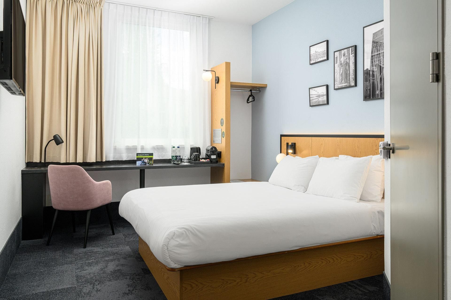 Quarto de hotel com cama, escrivaninha, cadeira, janela com cortinas e quadros na parede.