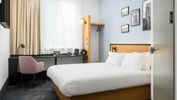 Quarto de hotel com cama, escrivaninha, cadeira, janela com cortinas e quadros na parede.