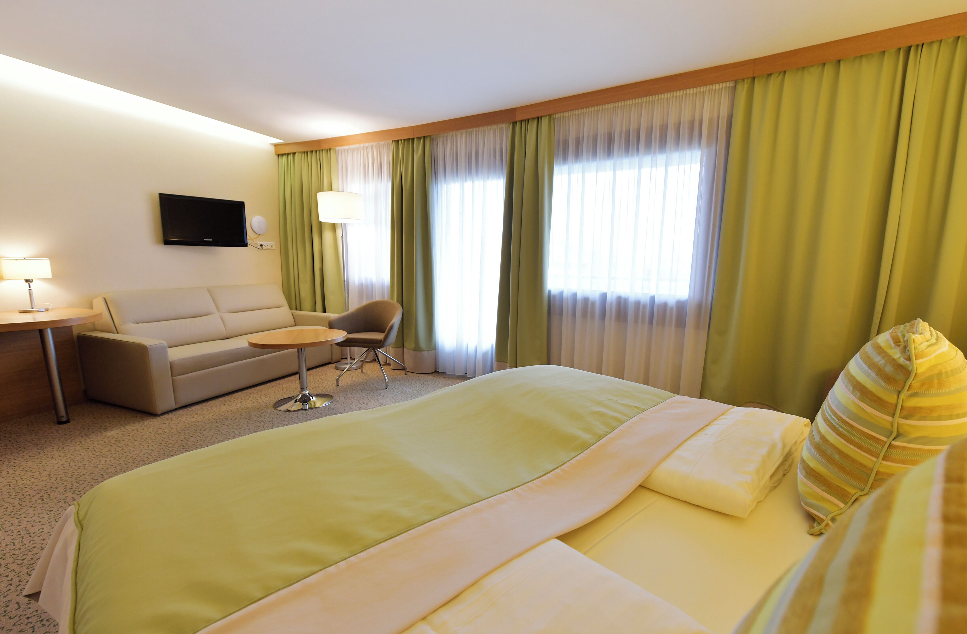 Quarto de hotel com cama, sofá, mesinha, televisão e cortinas.
