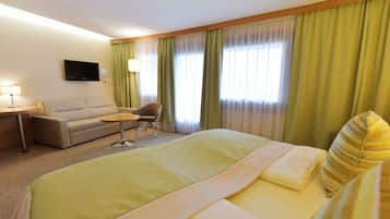 Quarto de hotel com cama, sofá, mesinha, televisão e cortinas.