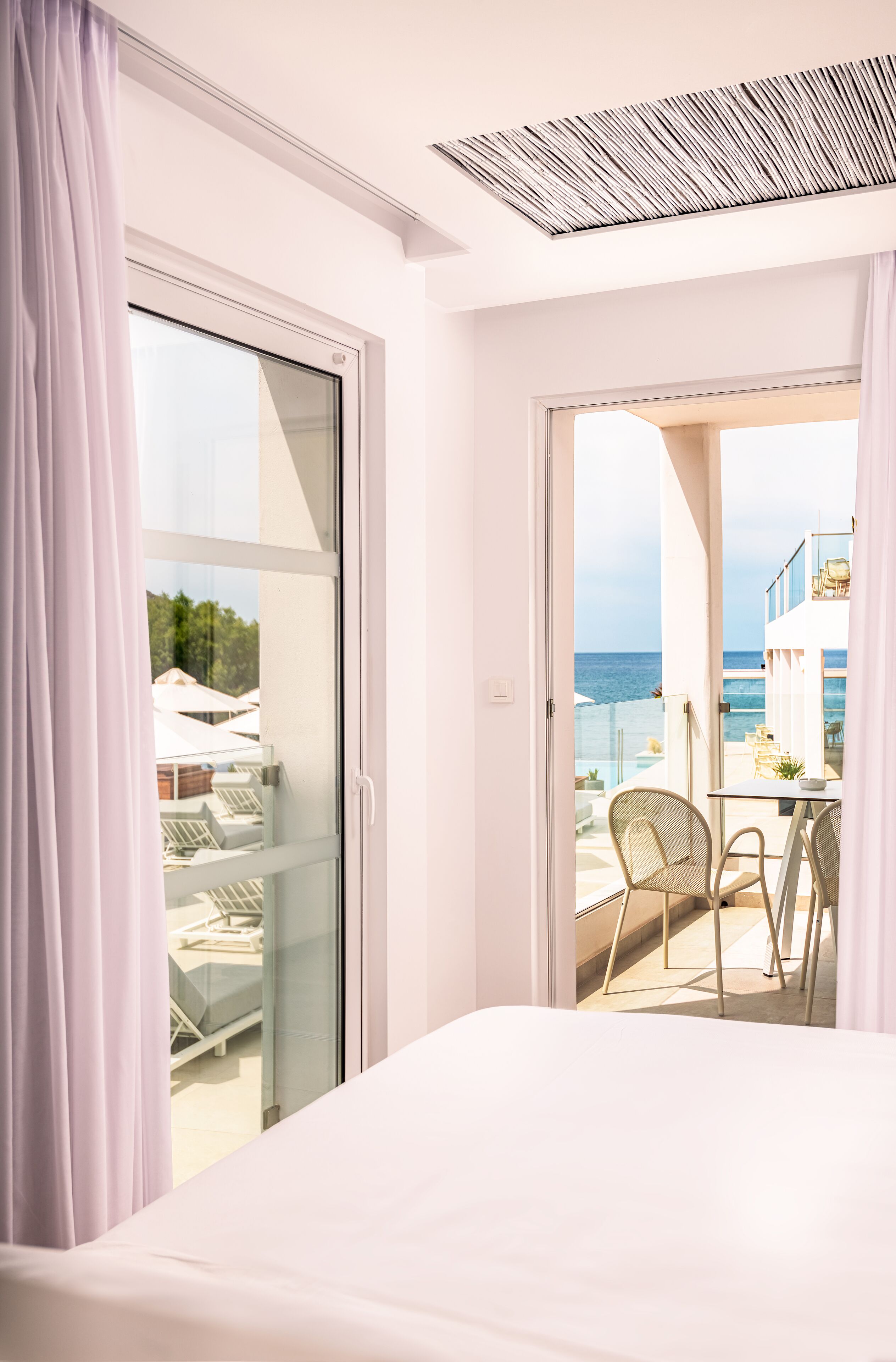 Quarto de hotel com uma cama grande, porta de vidro deslizante para uma varanda com mobiliário de exterior e vista para o mar.