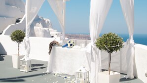 Um gazebo branco com tecido esvoaçante, uma mesa com toalha branca e duas pequenas plantas em vasos.