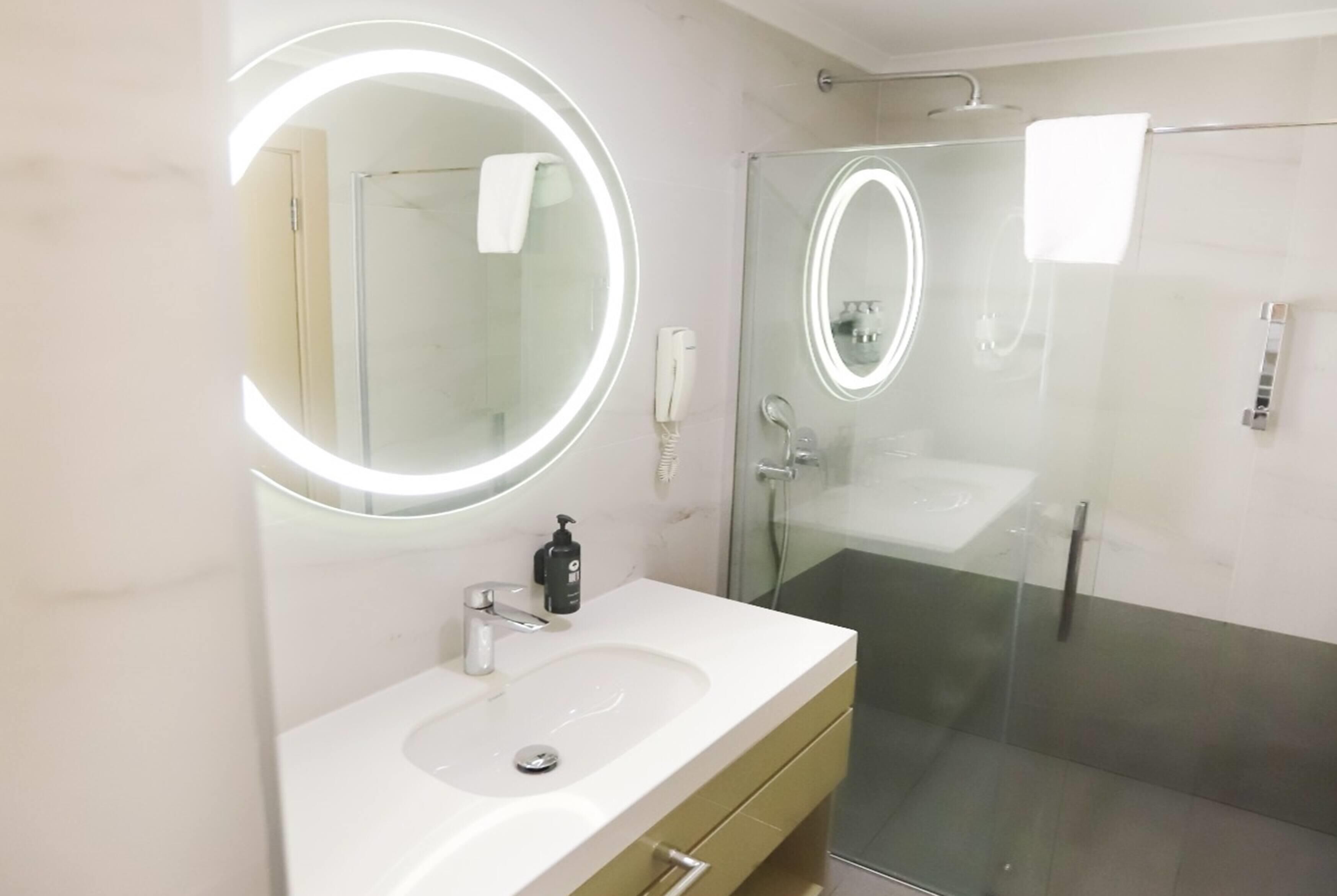Uma casa de banho moderna com um espelho redondo, lavatório, cabine de duche em vidro e chuveiro manual.