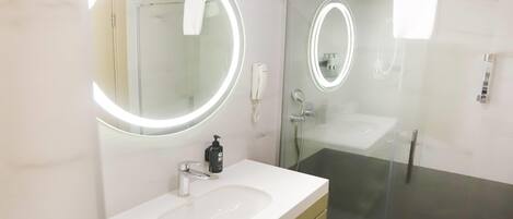 Uma casa de banho moderna com um espelho redondo, lavatório, cabine de duche em vidro e chuveiro manual.