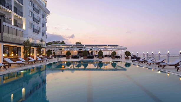 Área da piscina com espreguiçadeiras e um bar, cercada por um edifício com vários andares e varandas.