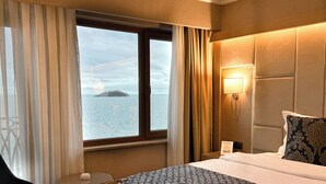Quarto de hotel com cama, uma cadeira, uma mesinha, janela com cortinas e uma luminária na parede.