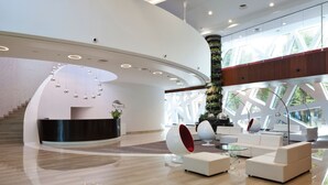 Um lobby moderno com um balcão de recepção curvo, área de estar com sofás brancos e uma escada com guarda-corpos de vidro.