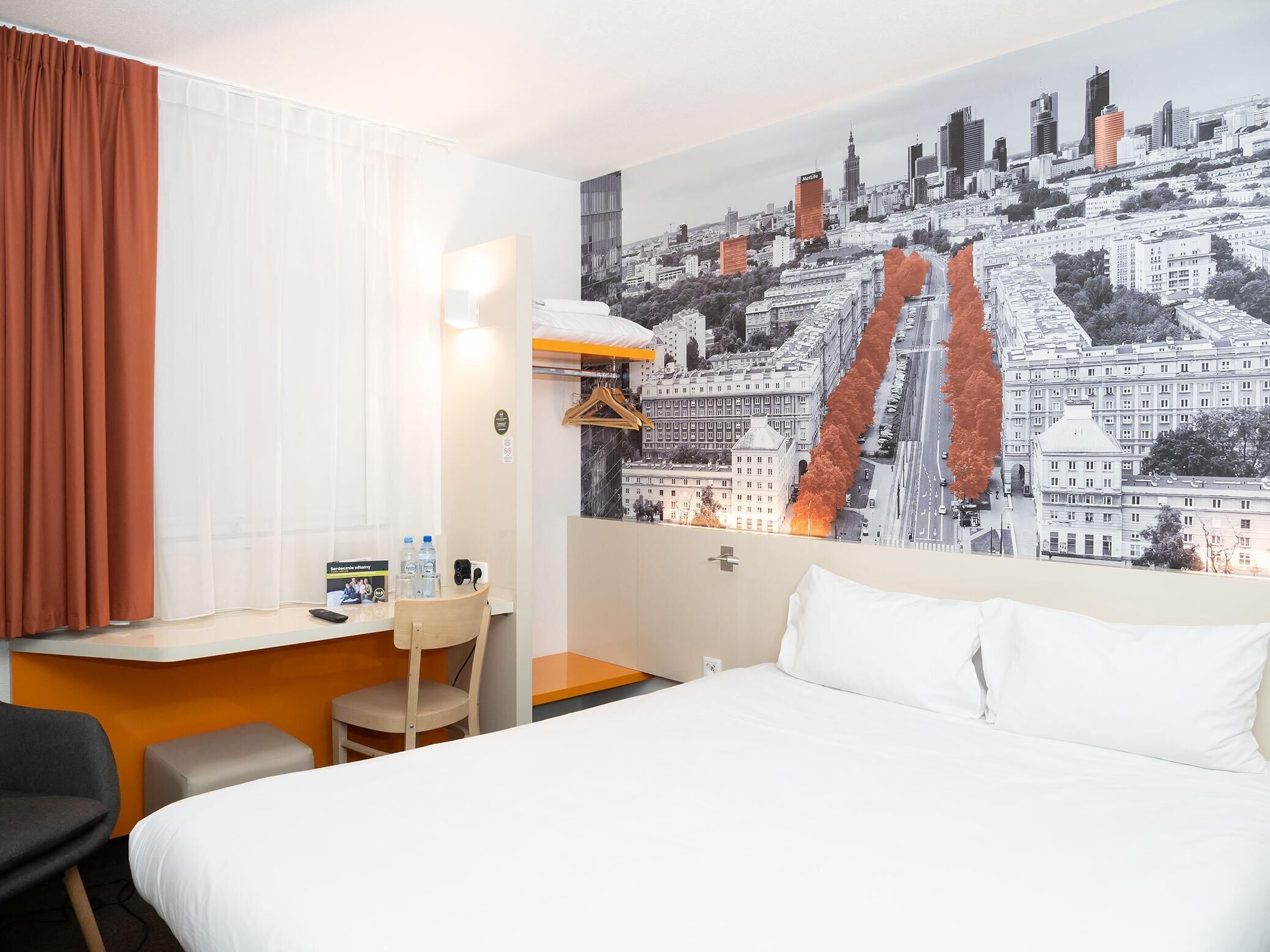 Quarto de hotel com cama, escrivaninha e cadeira, cabideiro e um mural com vista para a cidade na parede.