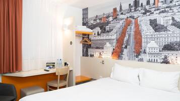 Quarto de hotel com cama, escrivaninha e cadeira, cabideiro e um mural com vista para a cidade na parede.