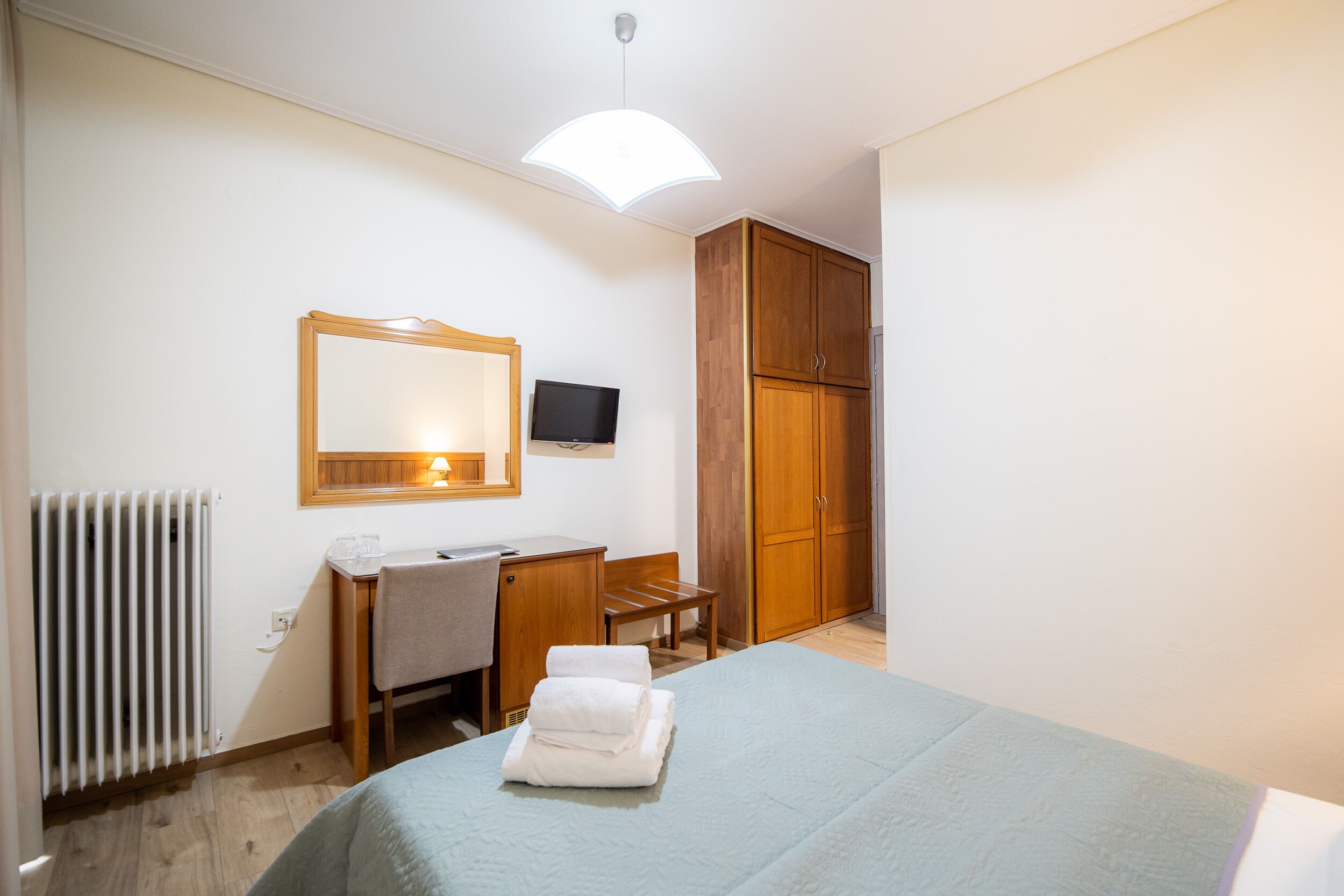 Quarto de hotel com cama, escrivaninha, cadeira, televisão e guarda-roupa.