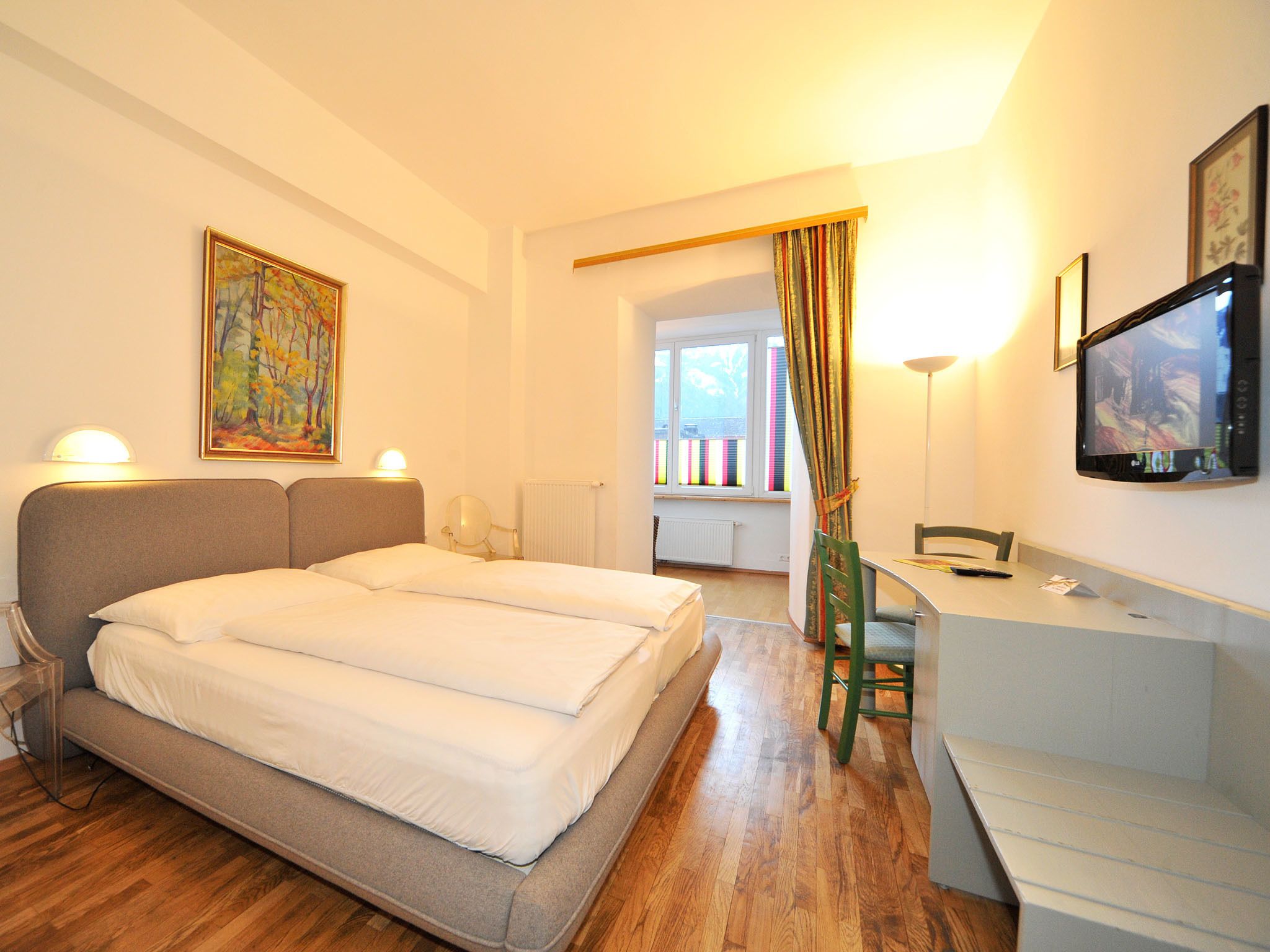 Quarto de hotel com cama, escrivaninha, cadeira, televisão, janela com cortinas e um quadro na parede.