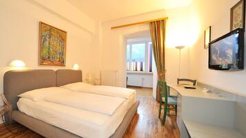 Quarto de hotel com cama, escrivaninha, cadeira, televisão, janela com cortinas e um quadro na parede.
