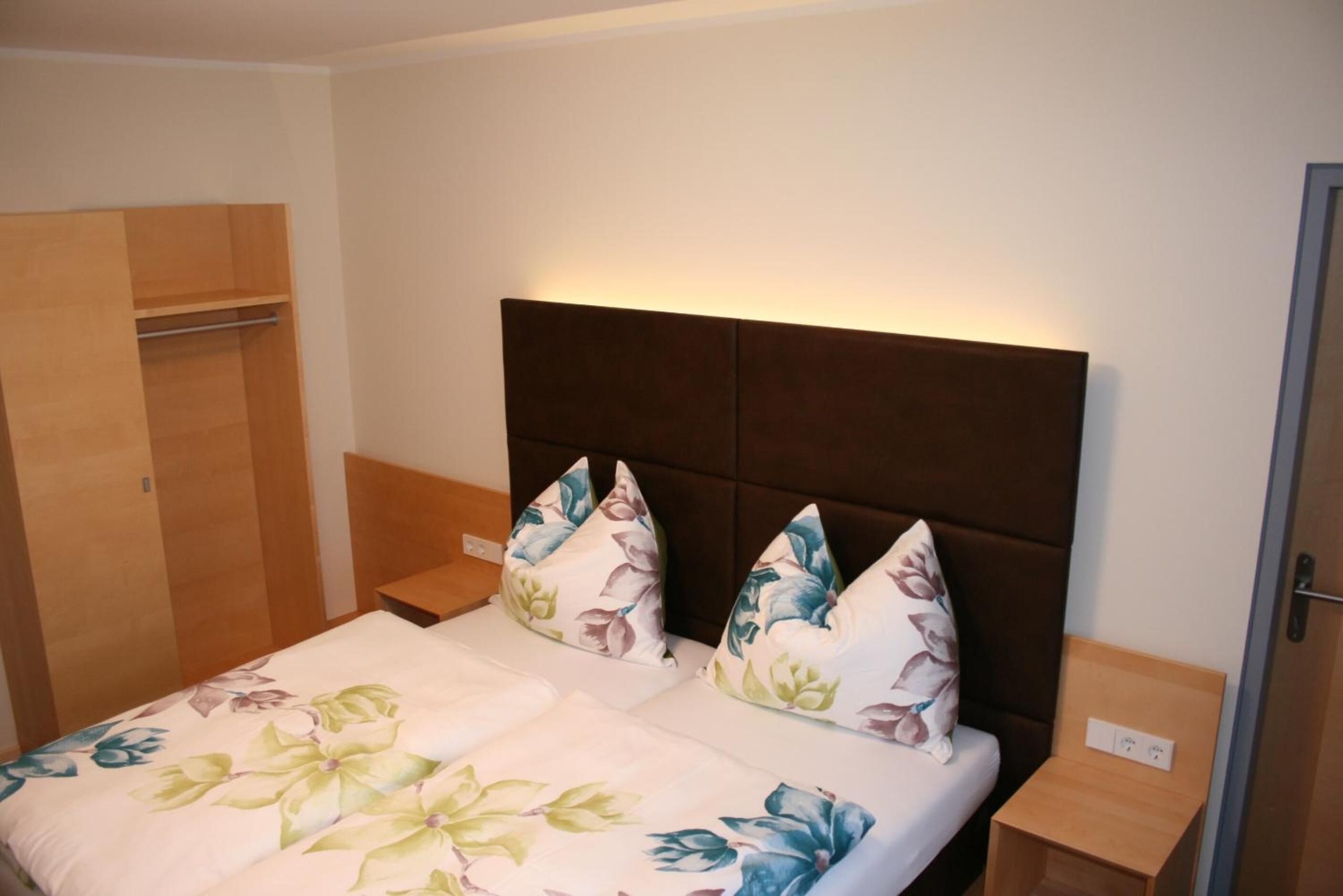 Quarto com cama, duas mesas de cabeceira, guarda-roupa de madeira e cabeceira com painel de luz.