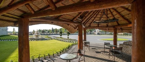 Um pátio externo com telhado de palha, área de estar e vista para um jardim ajardinado e estrada.