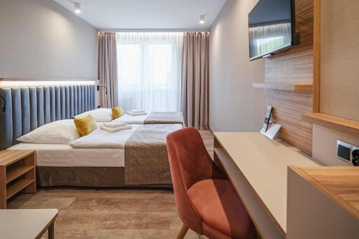 Quarto de hotel com cama, escrivaninha, cadeira, televisão e janela com cortinas.