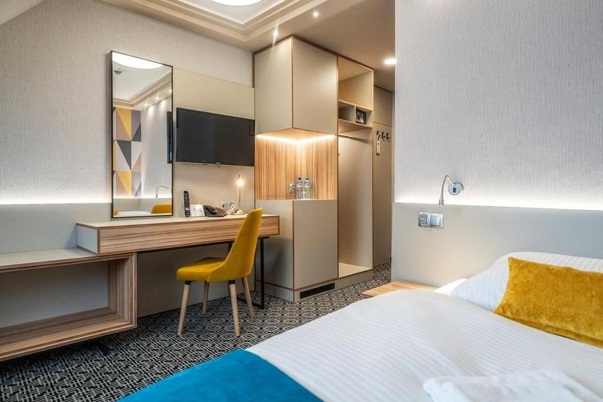 Quarto de hotel com uma cama, uma escrivaninha, uma cadeira, um espelho, uma televisão e um guarda-roupa.