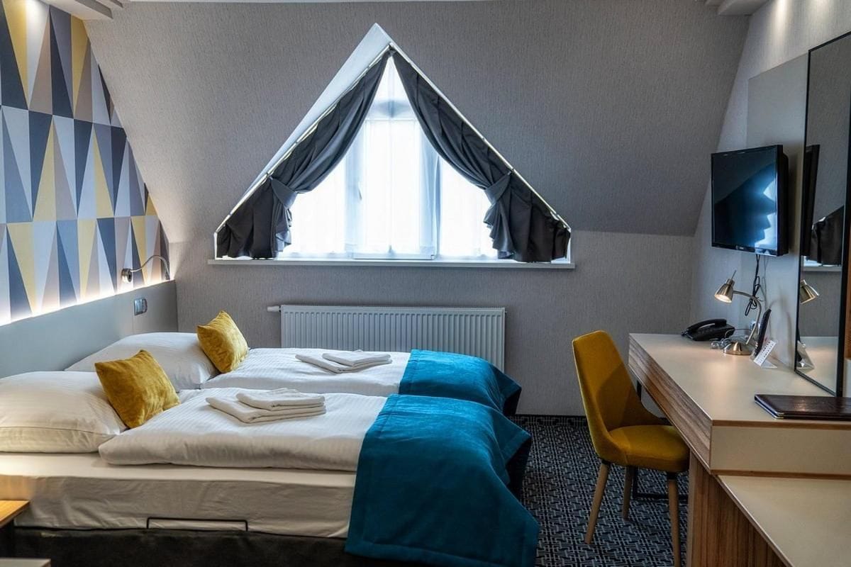 Quarto de hotel com uma cama, uma escrivaninha, uma cadeira, uma televisão e uma janela com cortinas.