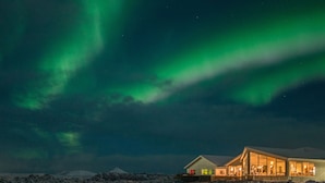 Casa com grandes janelas e paisagem coberta de neve sob um céu noturno com aurora verde.