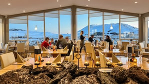 Restaurante com grandes janelas que oferecem vista para uma paisagem nevada e montanhas. Mesas montadas com velas e copos de vinho.