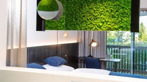 Quarto de hotel moderno com uma janela grande, cama com roupa de cama azul, escrivaninha com cadeira e parede verde.