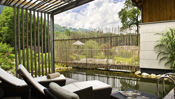 Área da piscina com espreguiçadeiras e uma pequena mesa, cercada por cercas de bambu e um deck de madeira.