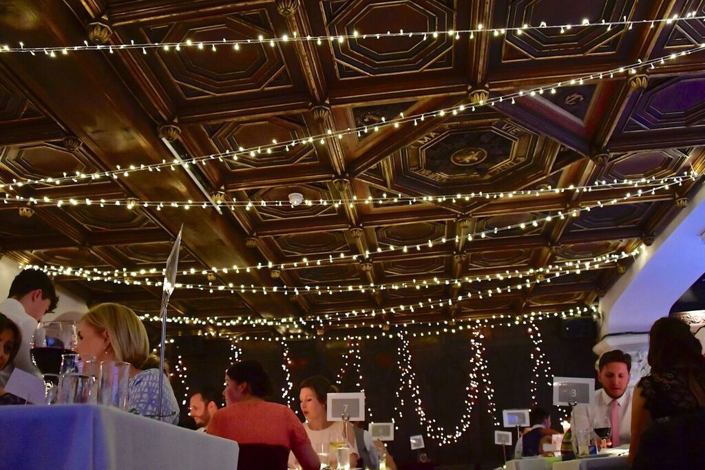 Restaurante com teto de madeira e luzes de corda. Mesas montadas com copos e pratos.