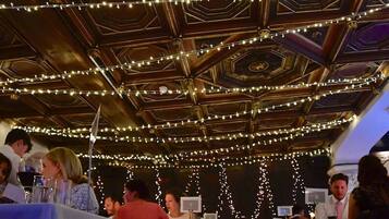 Restaurante com teto de madeira e luzes de corda. Mesas montadas com copos e pratos.