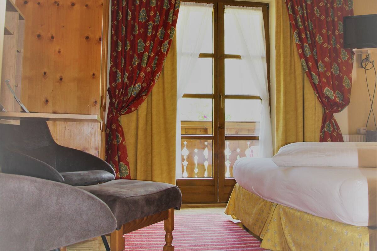Quarto com cama, escrivaninha e cadeira. Janela com cortinas e vista para o exterior.