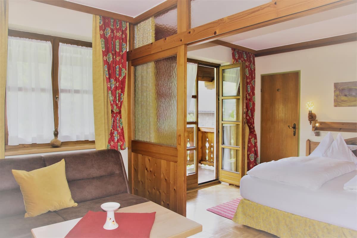 Quarto com cama de madeira, sofá com almofada amarela, mesa pequena com guardanapo vermelho e porta que dá para outro cômodo.