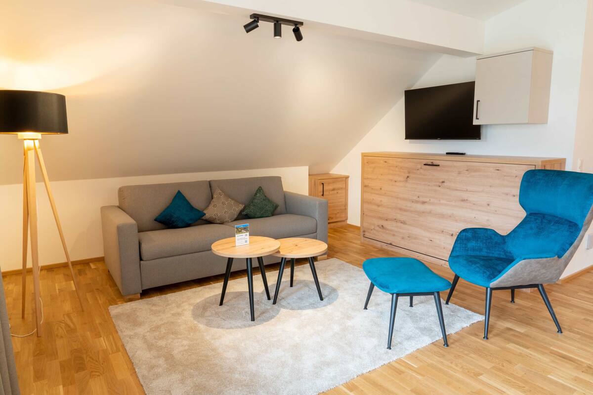 Uma sala de estar moderna com um sofá cinzento, duas mesas redondas, uma poltrona azul, um armário de madeira e uma TV de tela plana.