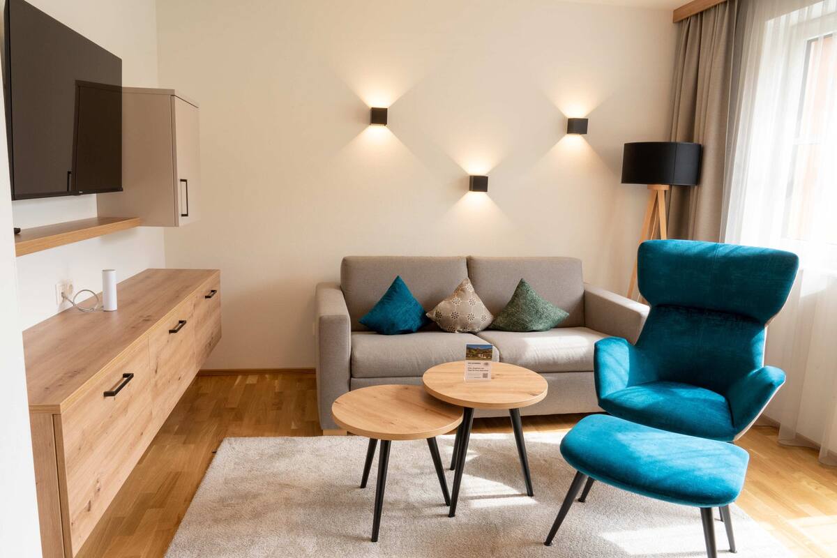 Uma sala de estar moderna com um sofá cinza, uma poltrona azul, uma mesa lateral de madeira e uma pequena mesa redonda.