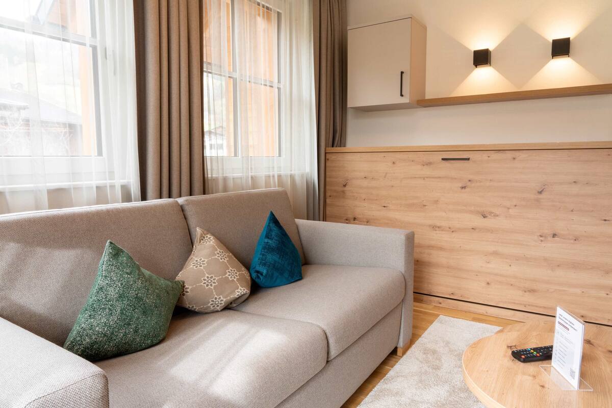 Uma sala de estar moderna com um sofá bege, almofadas estampadas, um aparador de madeira e um controle remoto sobre uma mesinha.
