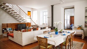Uma sala de estar ampla, com uma mesa de jantar posta para quatro pessoas, um sofá, uma escada e uma televisão sobre um móvel de madeira.