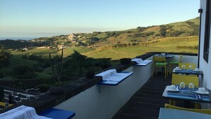 Um terraço com mesas e cadeiras, com vista para uma paisagem rural com campos e uma aldeia ao longe.