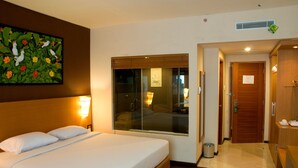 Quarto de hotel com uma cama grande, uma janela com persianas, uma obra de arte na parede, um abajur e uma porta de madeira que dá para um banheiro.