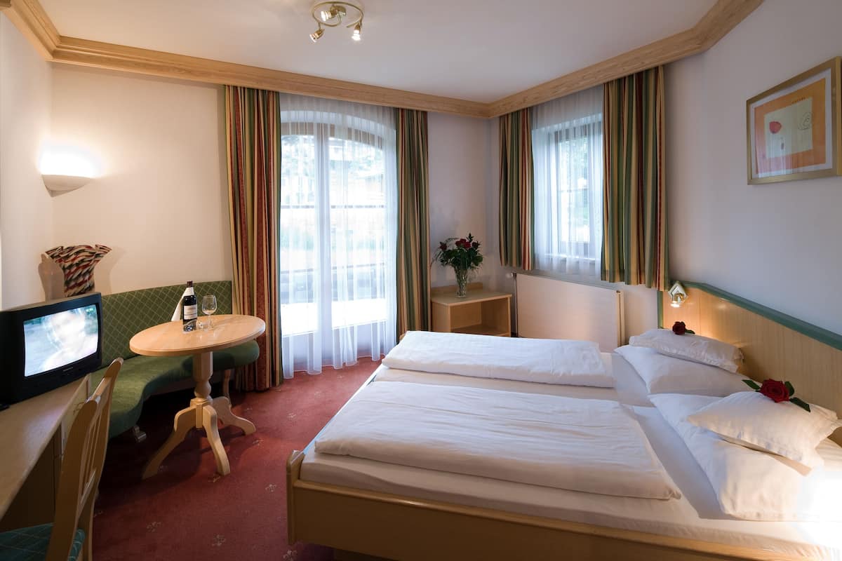 Quarto de hotel com duas camas, uma mesa pequena com garrafa e copo, uma cadeira, televisão e janela com cortinas.