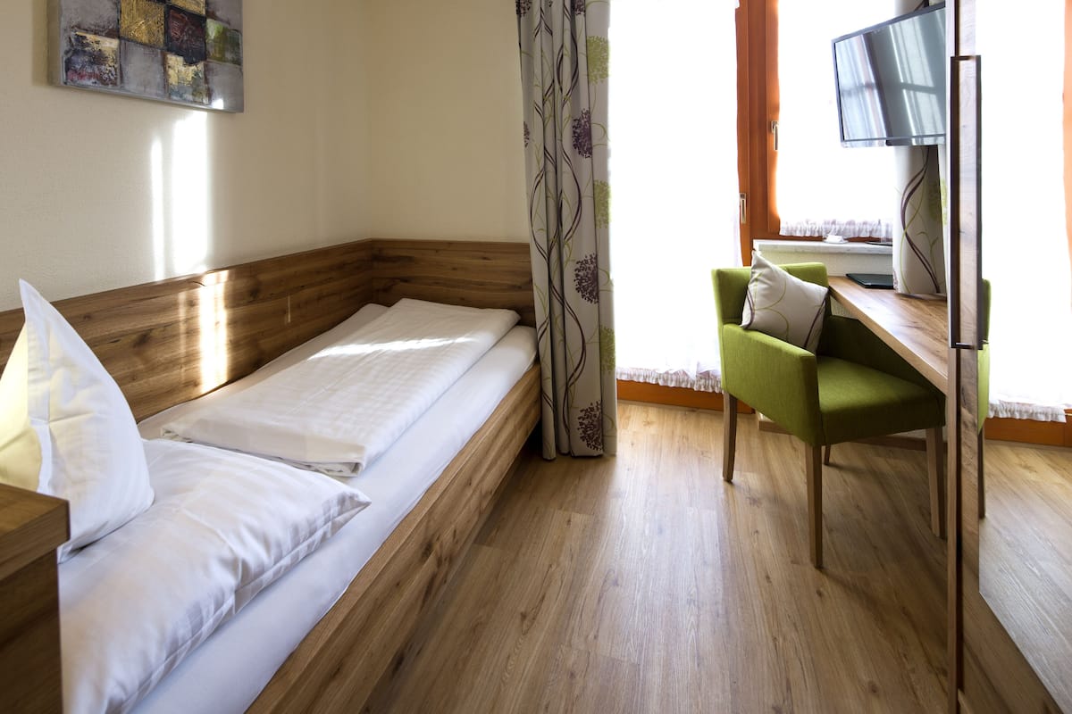 Quarto com cama de madeira, uma cadeira verde, escrivaninha com televisão e janela com cortinas.