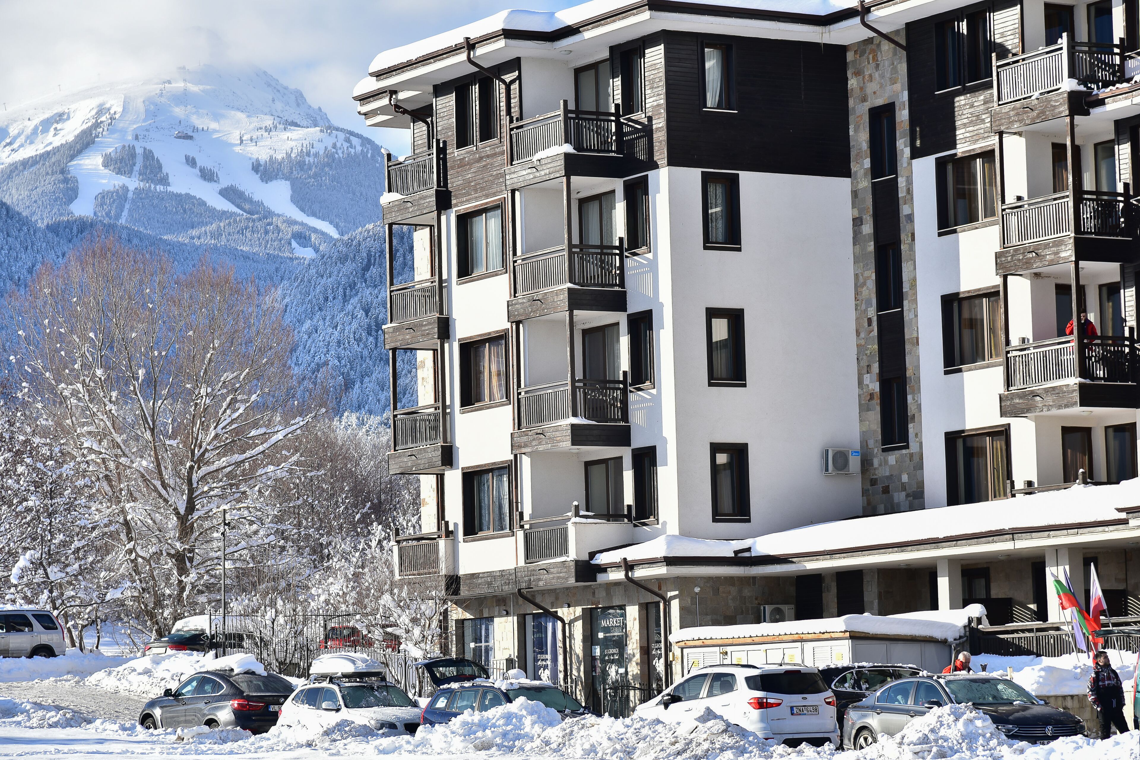 Um hotel com vários andares e varandas, um estacionamento coberto de neve e uma montanha ao fundo.
