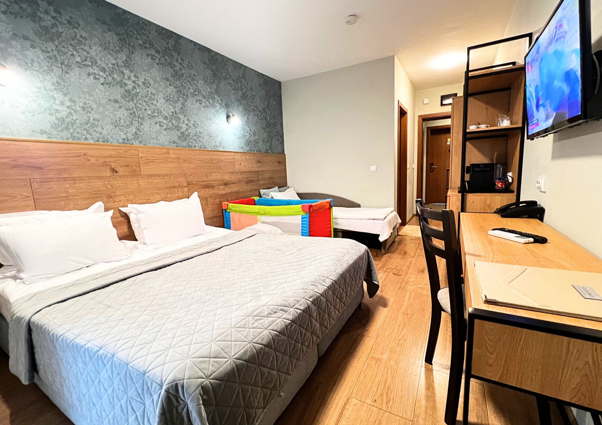 Quarto de hotel com uma cama, escrivaninha, cadeira, televisão e guarda-roupa.