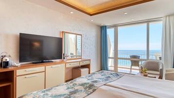 Quarto de hotel com cama, televisão, escrivaninha e vista para o mar.