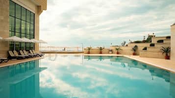 Piscina com espreguiçadeiras e guarda-sóis, cercada por edifícios e plantas em vasos.