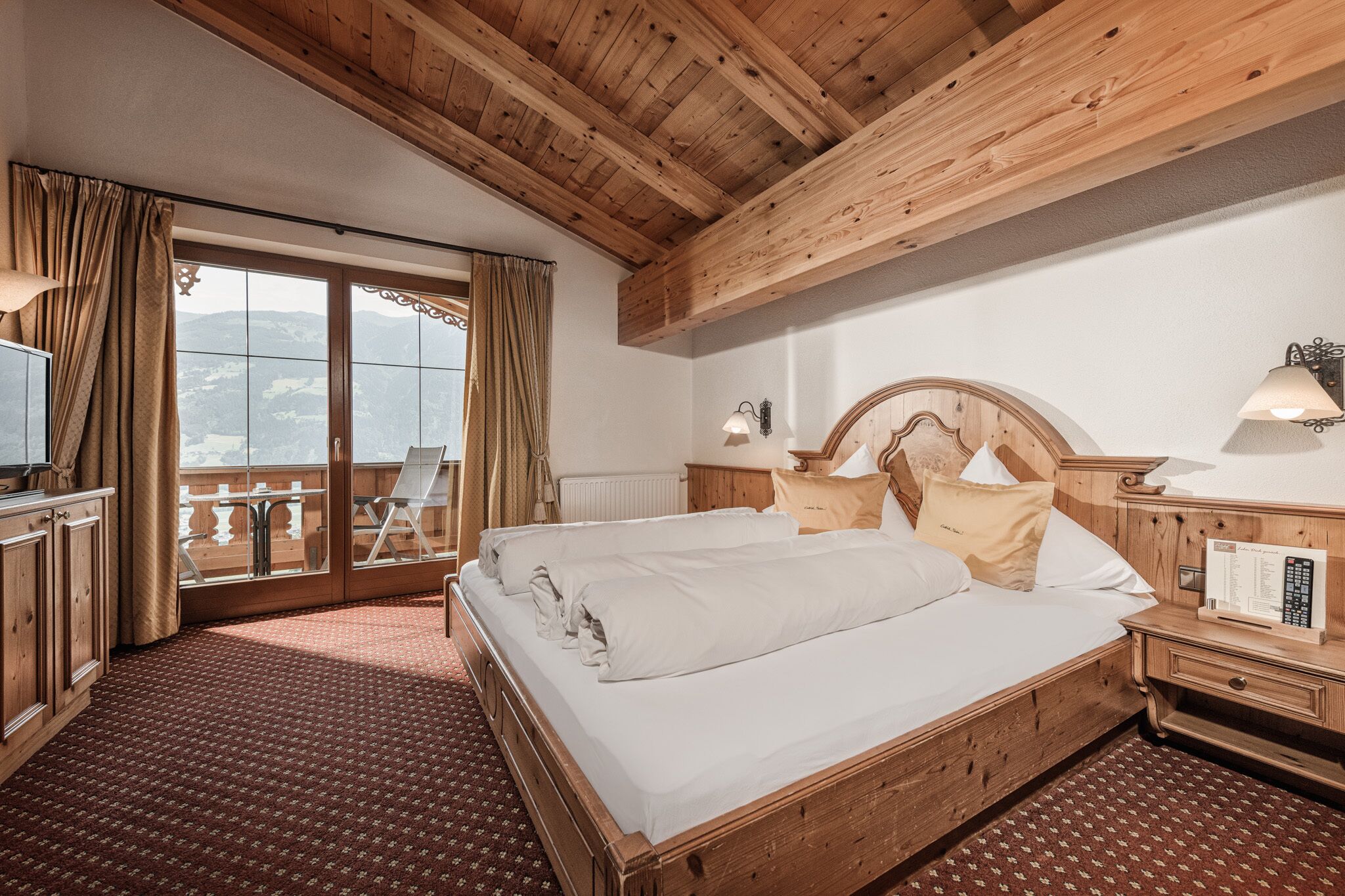 Uma cama de madeira com roupa de cama branca, uma mesa de cabeceira de madeira com uma luminária e vista para montanhas através de uma grande janela.