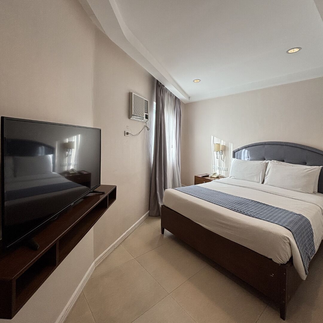 Quarto de hotel com uma TV de tela plana grande sobre um suporte de madeira, uma cama com cabeceira escura, uma mesa de cabeceira com abajur e um ar-condicionado fixado na parede.