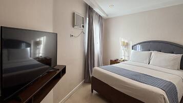 Quarto de hotel com uma TV de tela plana grande sobre um suporte de madeira, uma cama com cabeceira escura, uma mesa de cabeceira com abajur e um ar-condicionado fixado na parede.
